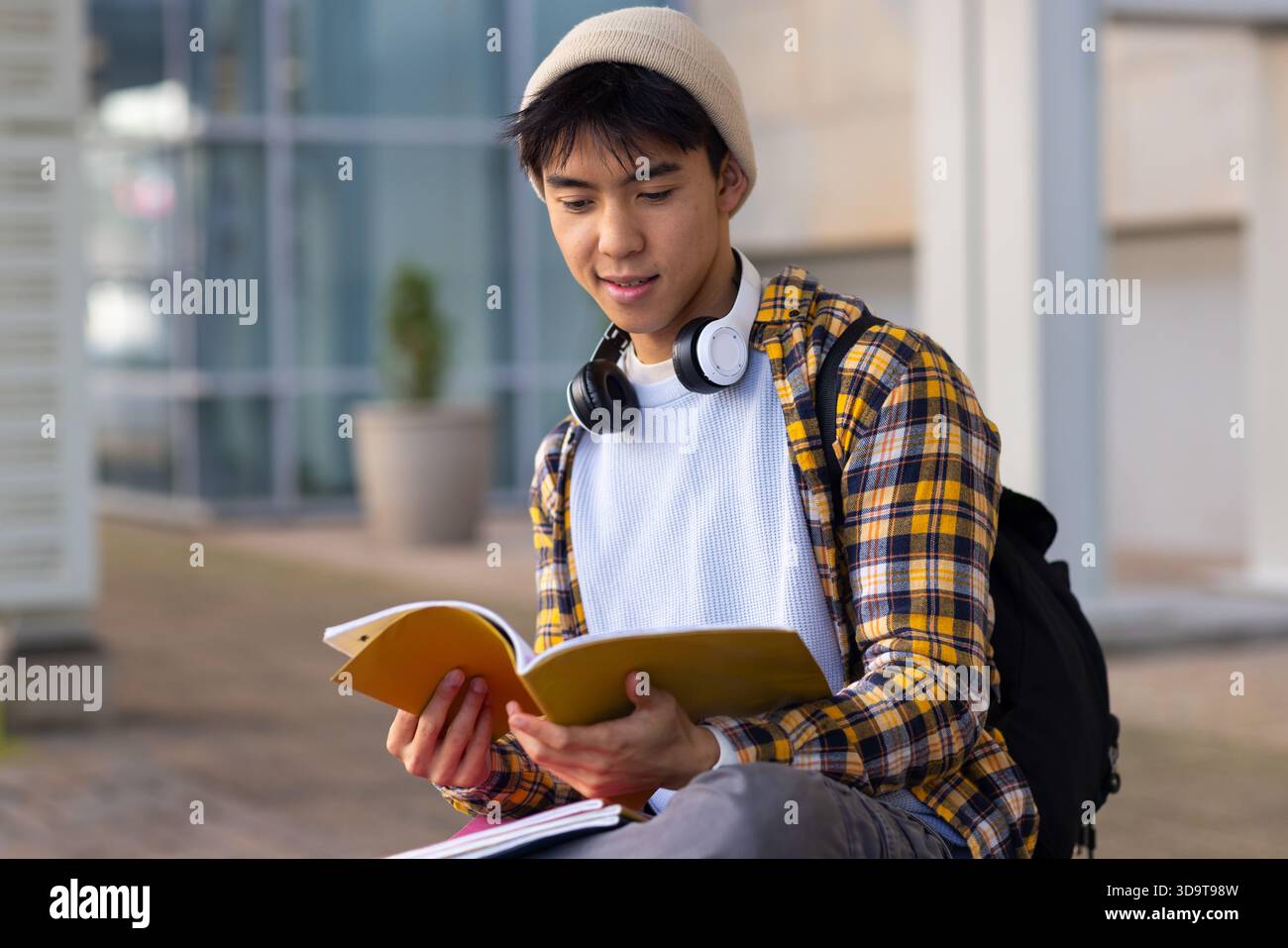 Asian male student sitting on ledge outside campus wearing headphones ...