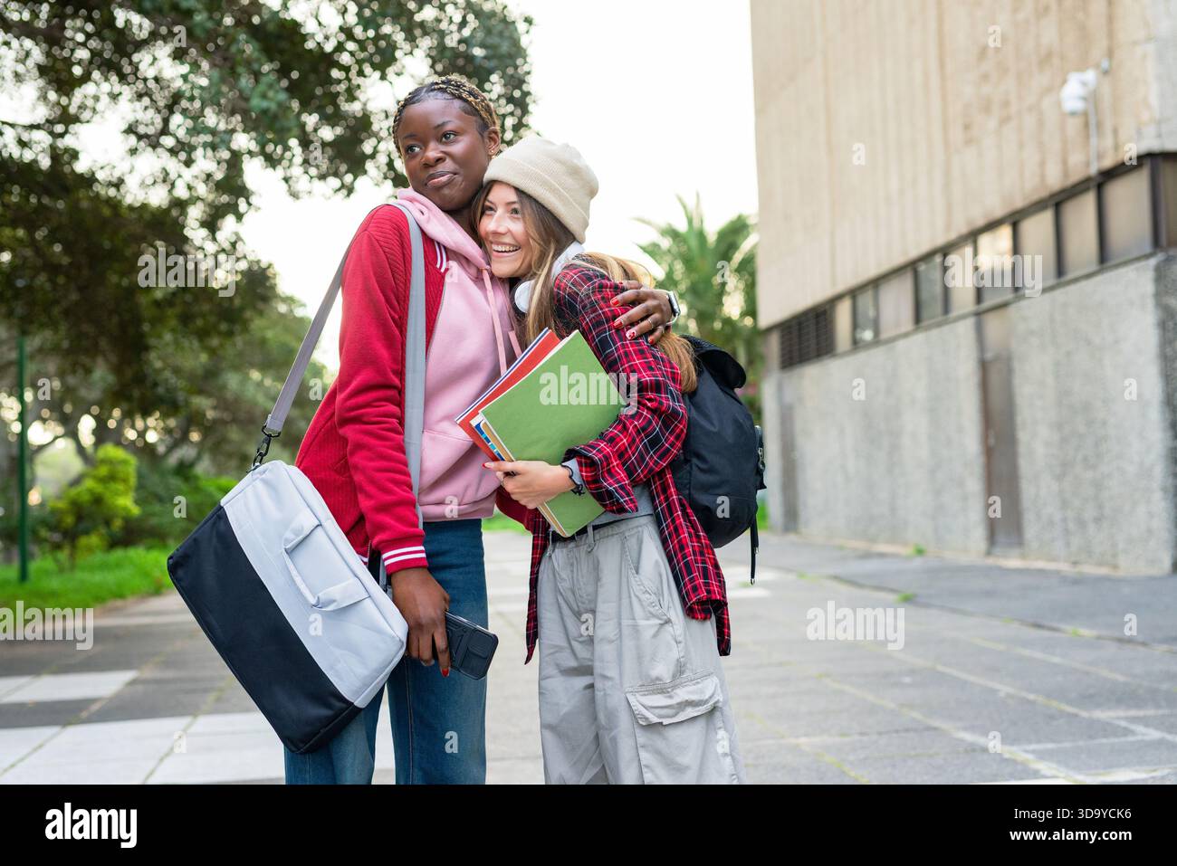 Diverse female students hugging on campus pathway carrying messenger ...