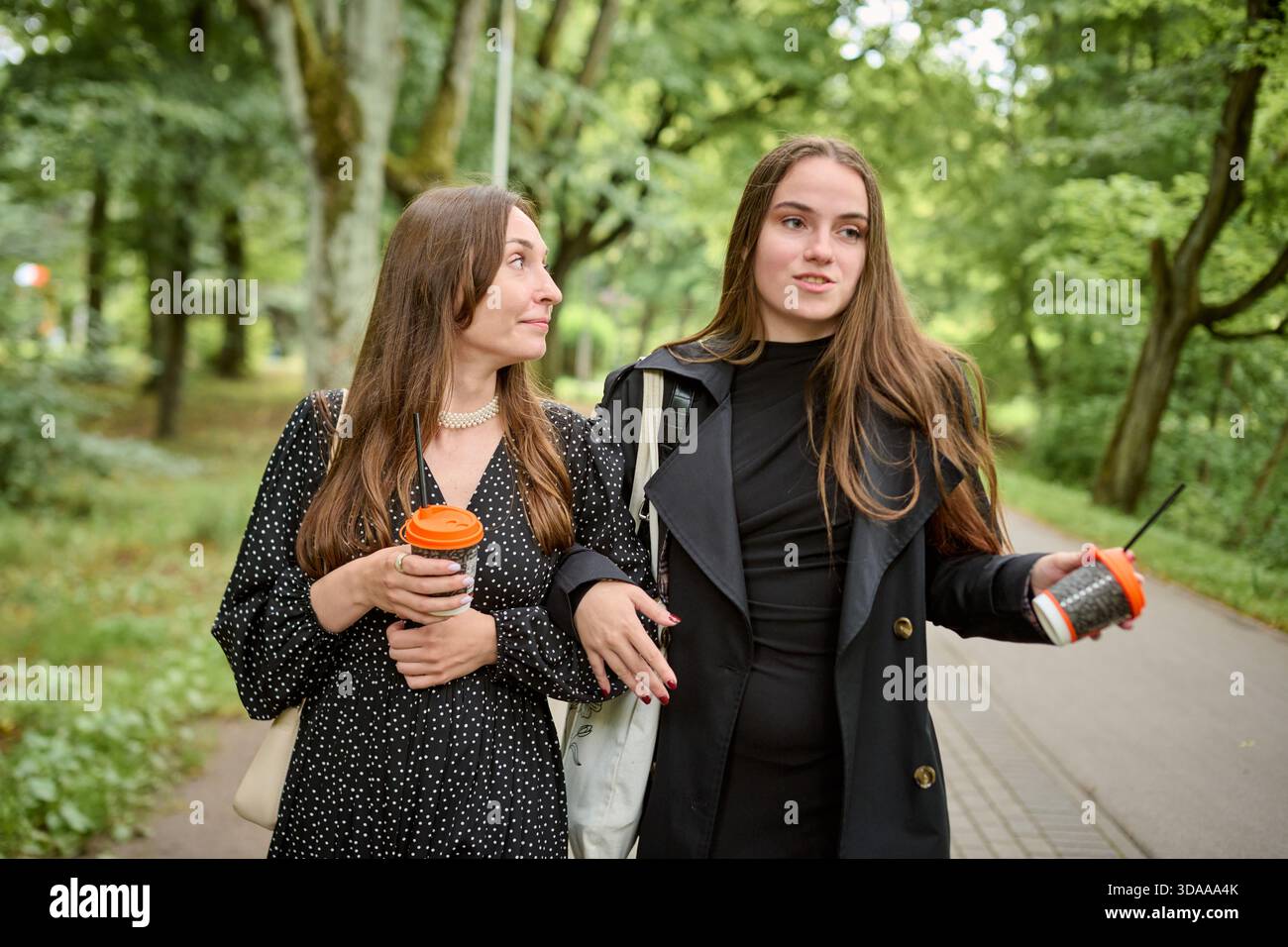 Two young women with long brown hair walk arm - In - Arm along shaded ...