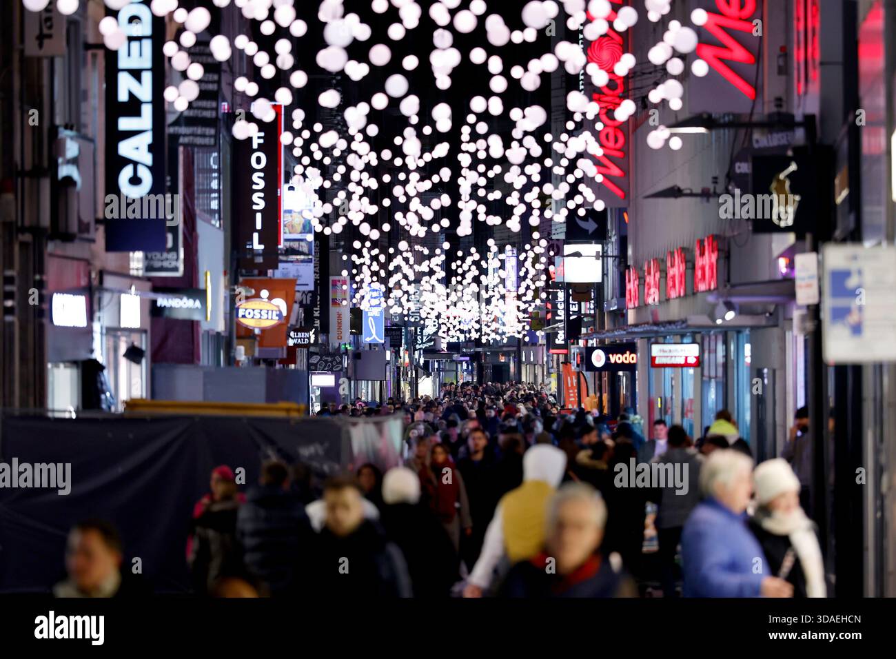 Passersby in the High Street pass by illuminated shops. The business ...