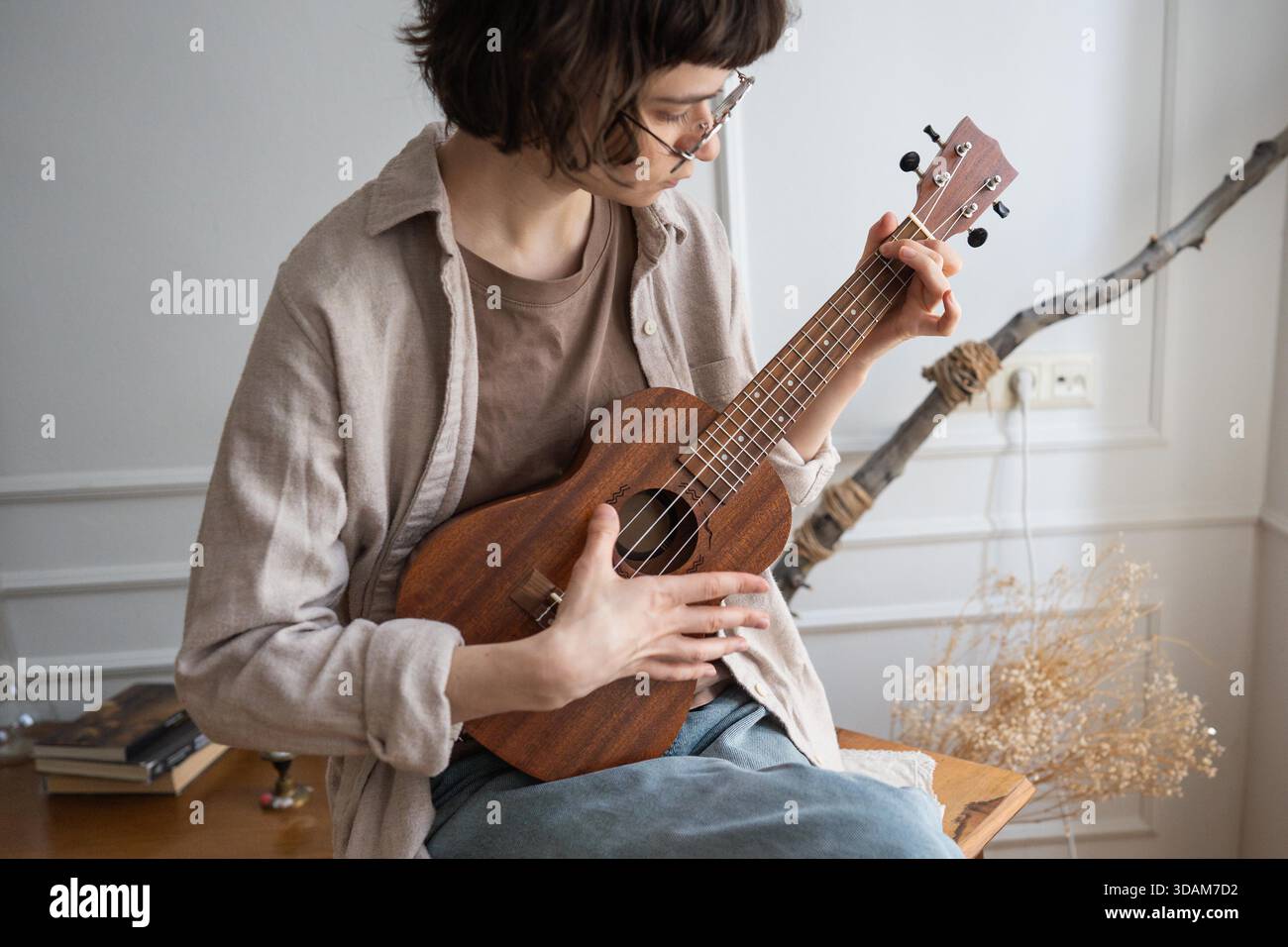 Focused young woman in glasses learning ukulele chords at home, intent ...