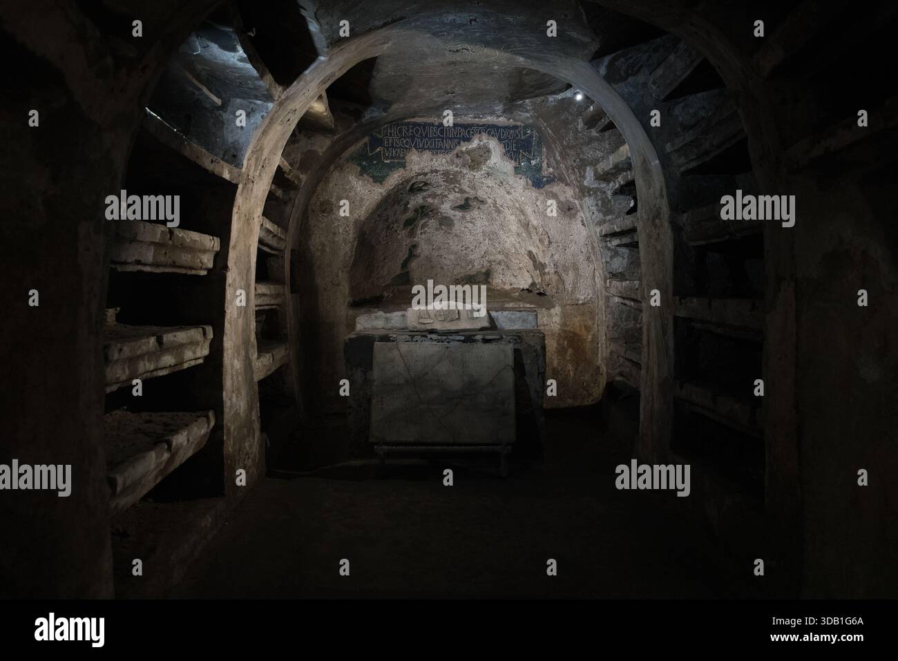 Dimly lit ancient catacomb chamber with stone shelves, altar and fresco ...