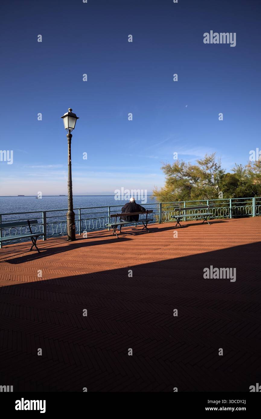 Elder man sitting on a bench by the edge of a promenade by the sea next ...