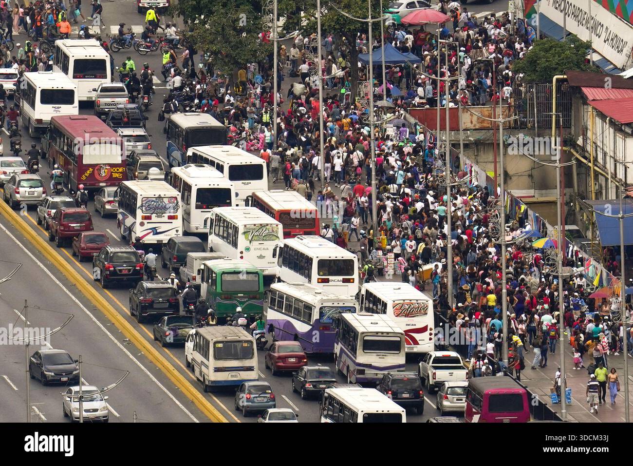 Market vendors and shoppers fill the side of a street as cars sit in ...