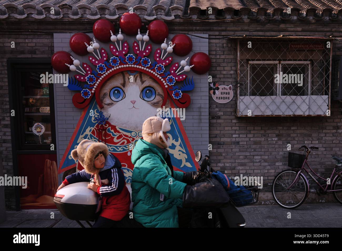 A man rides an electric bike with a child passing a cat drawing decorated with Peking Opera on ...