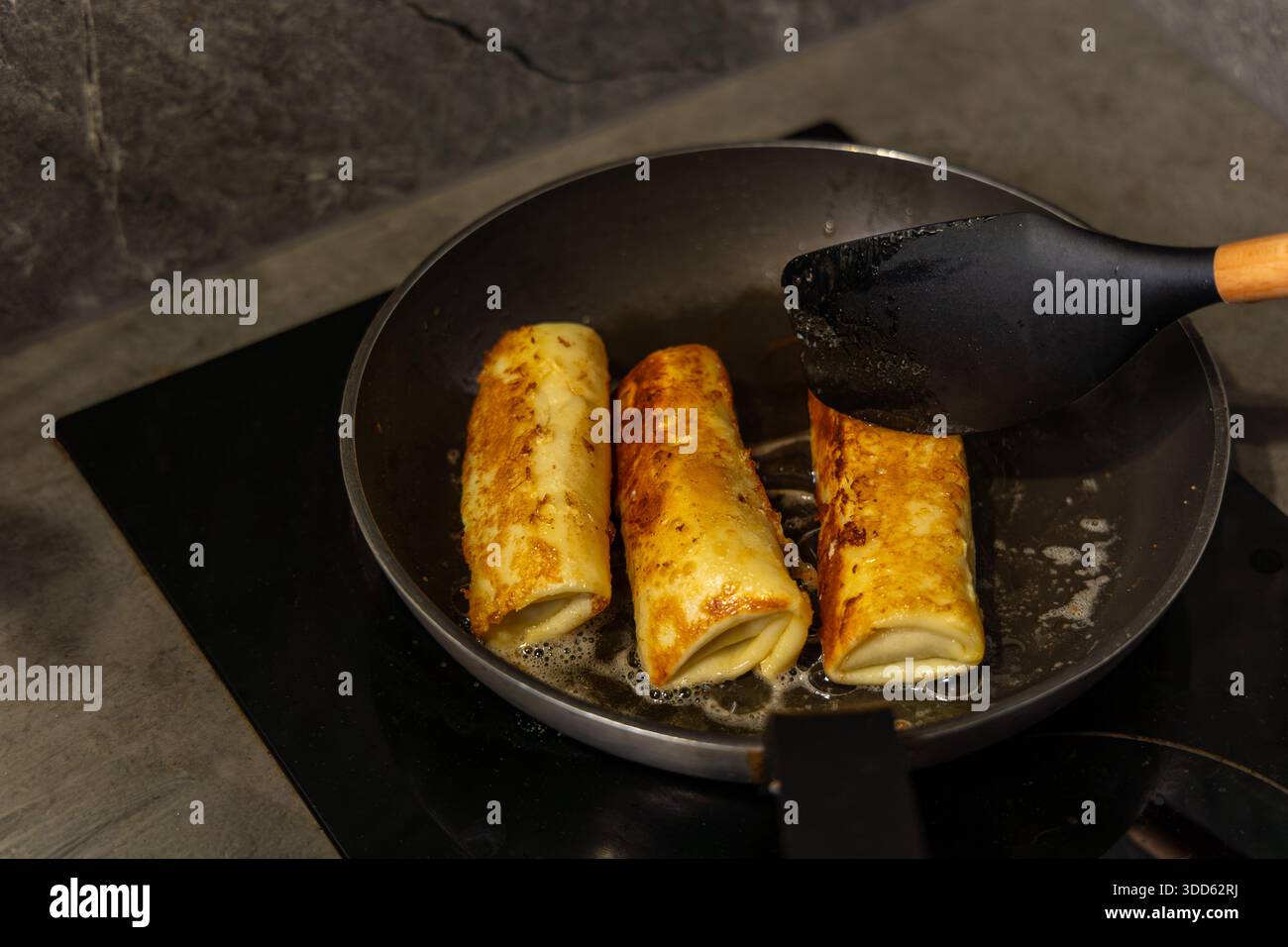 Golden crepes frying in pan on stove top. Three rolled crepes being pan ...