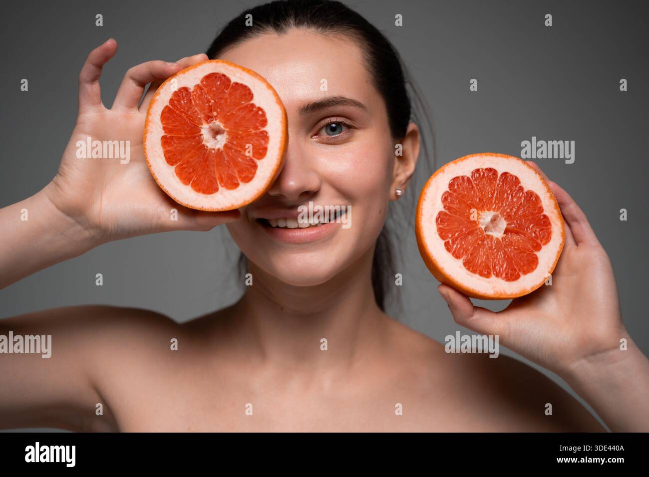 Smiling woman holding fresh grapefruit halves near face on neutral ...