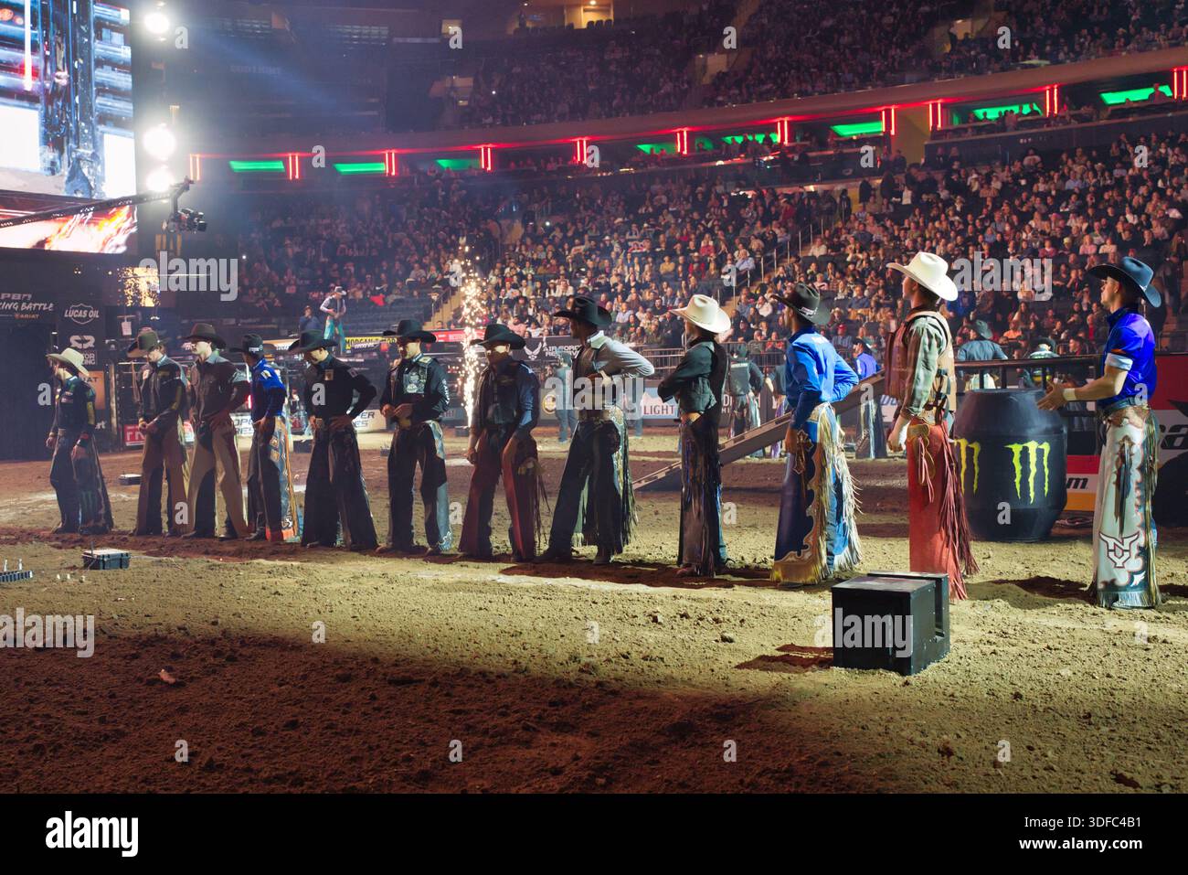 Cowboys stand in an arena before a large crowd. They wear traditional ...