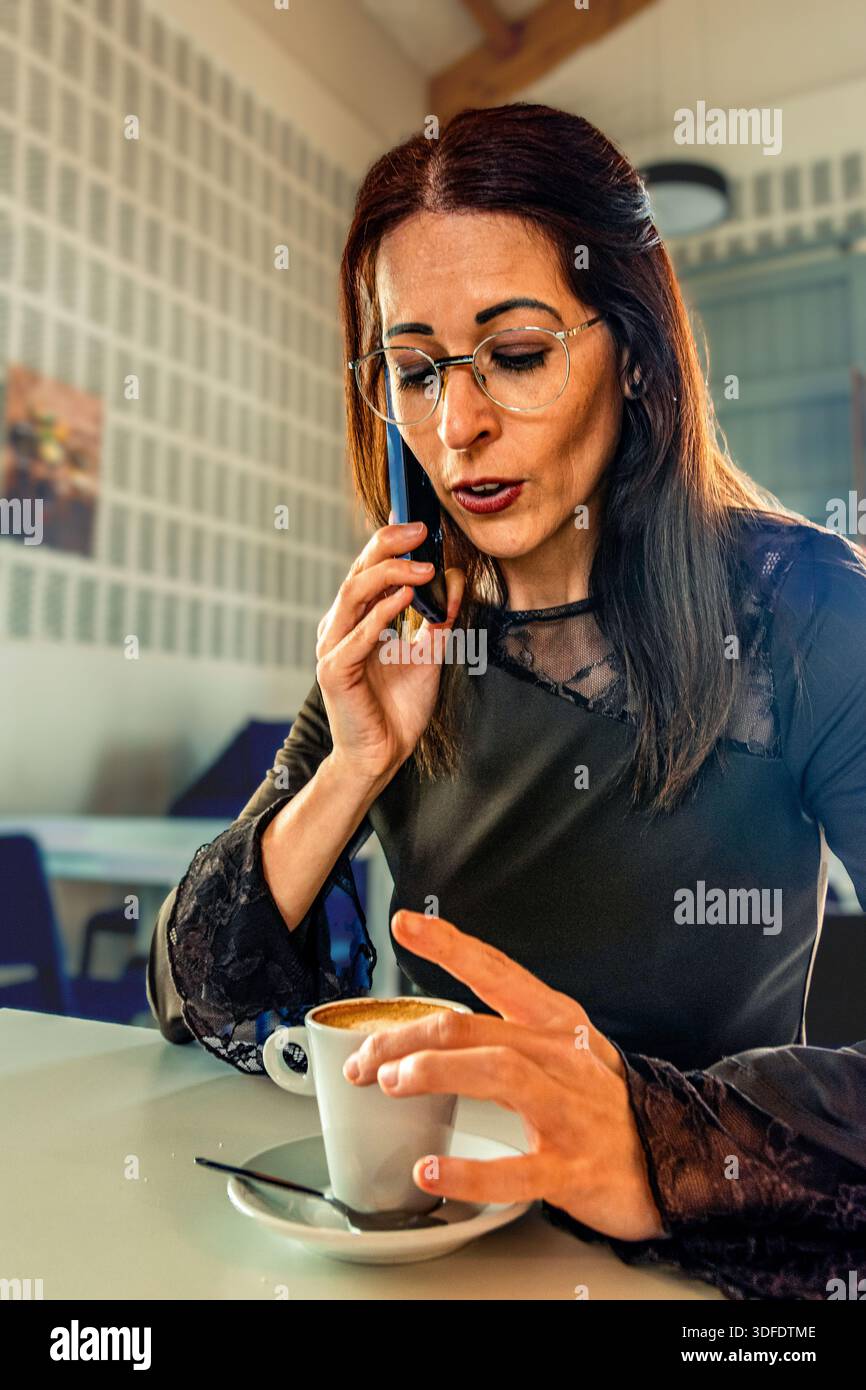 Woman in eyeglasses making a phone call while enjoying a cappuccino at ...