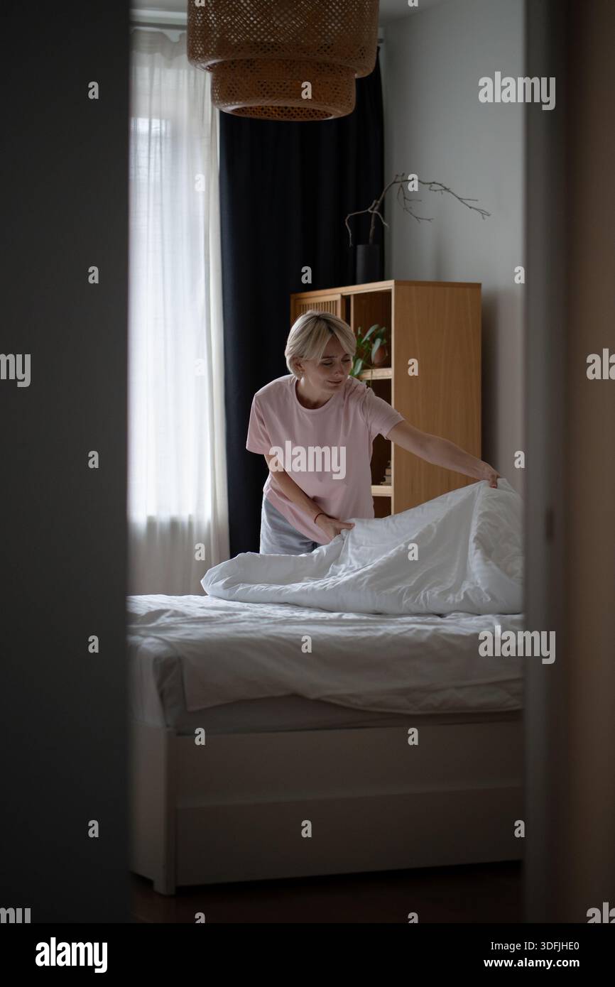 Relaxed smiling woman in bedroom tidying bed after night sleep ...