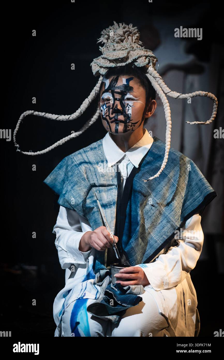 A performance artist sits under dramatic stage light, wearing an ethnic ...