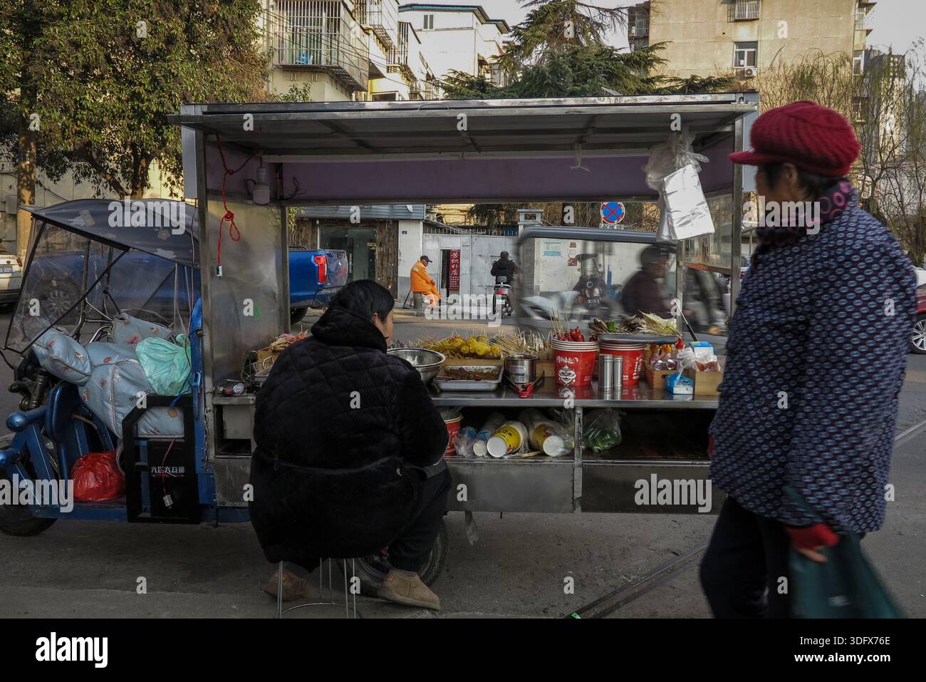 A street food cart sells skewers and snacks; a vendor sits by it, and a ...