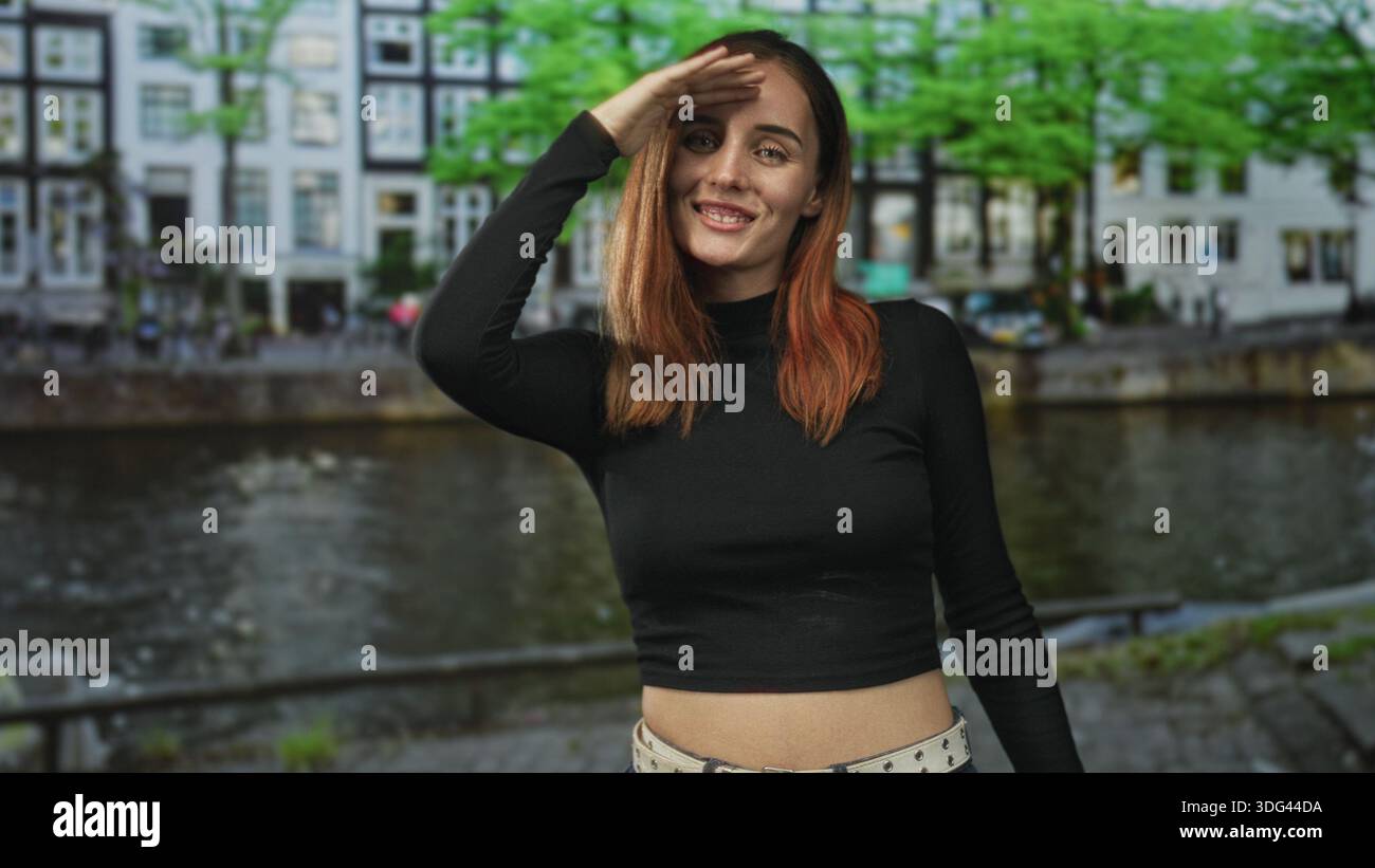 Woman showing midriff with hand shading eyes on street by a canal and ...
