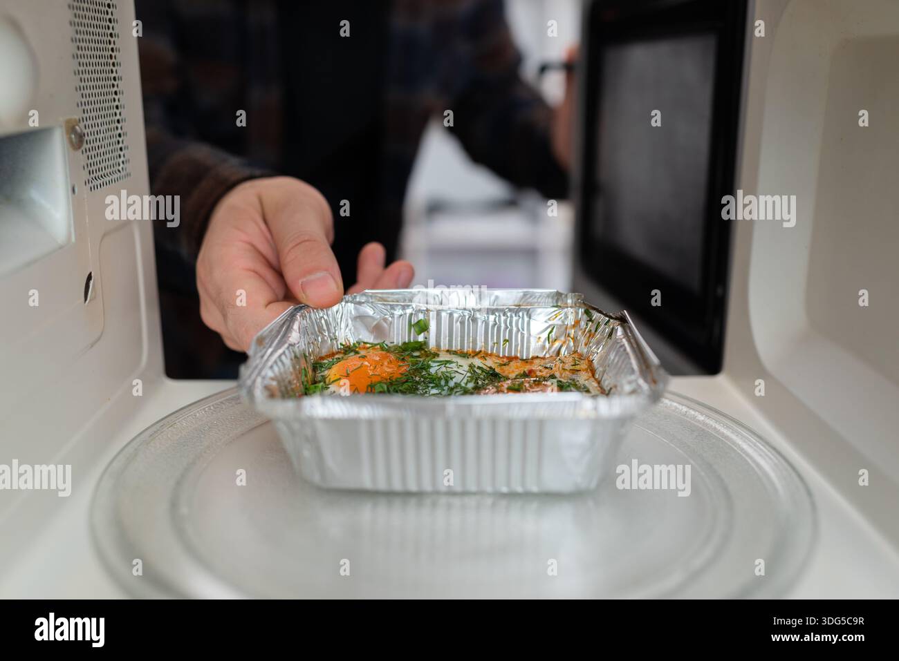 Man putting a foil container with fried eggs and herbs into a microwave ...