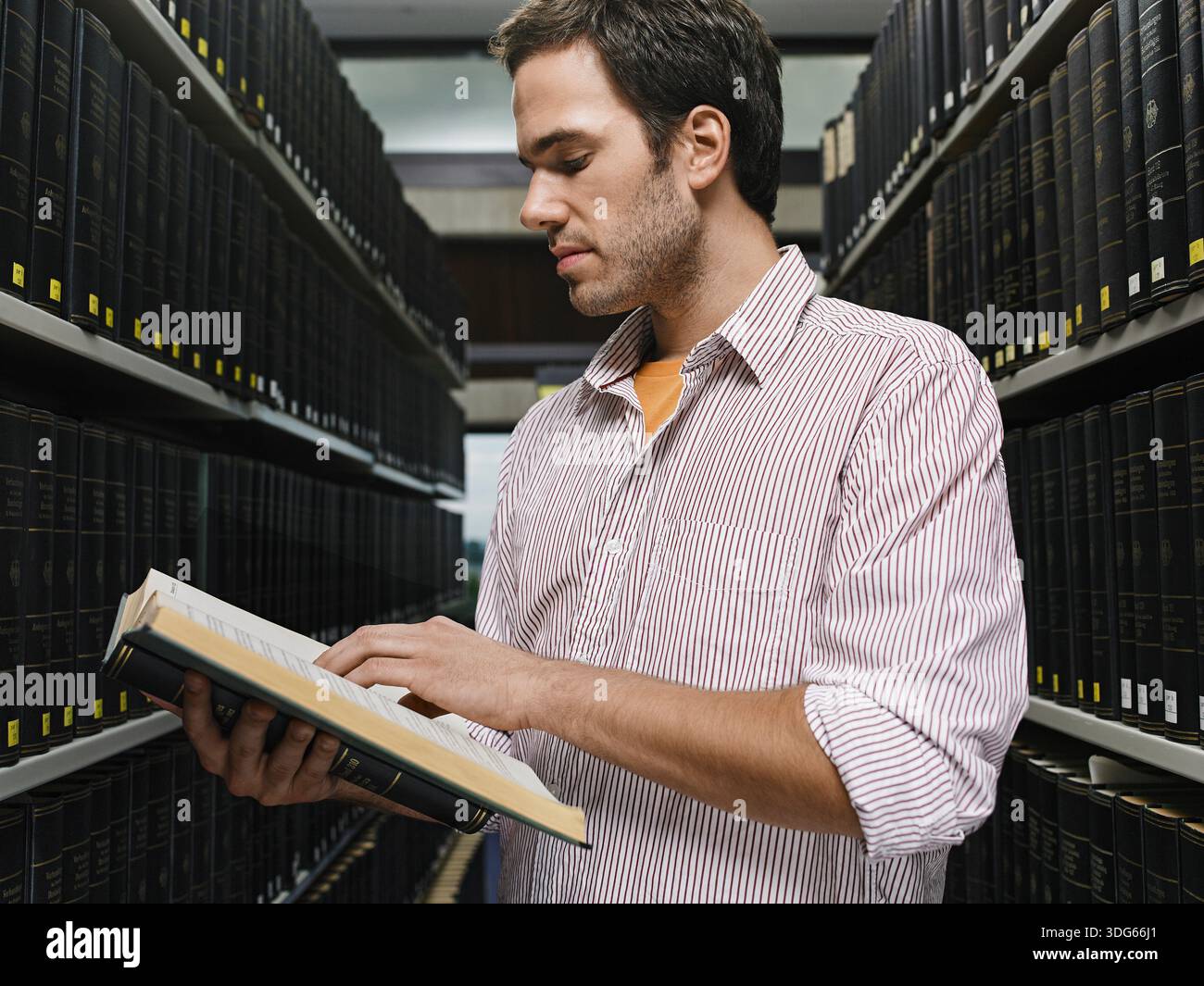 A young adult reading a book in a library aisle surrounded by shelves ...