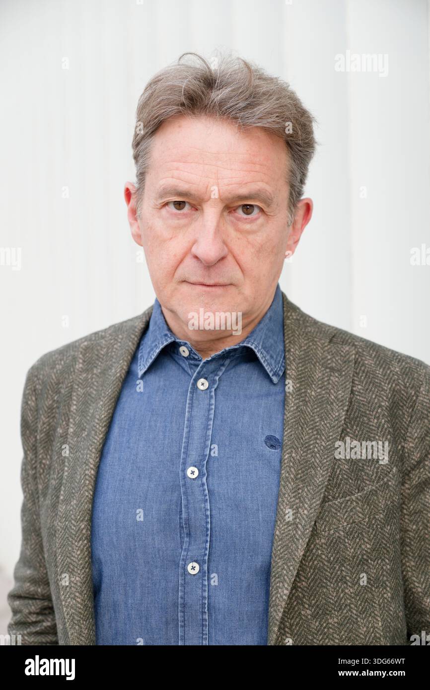 Man in a tweed jacket and denim shirt stands against a white wall with ...