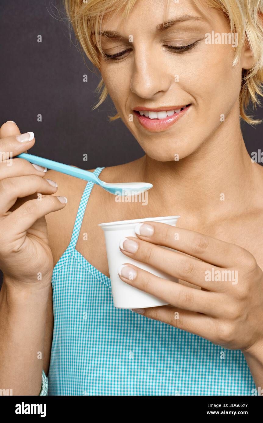 Smiling woman in a blue top enjoying yogurt with a spoon against a dark ...