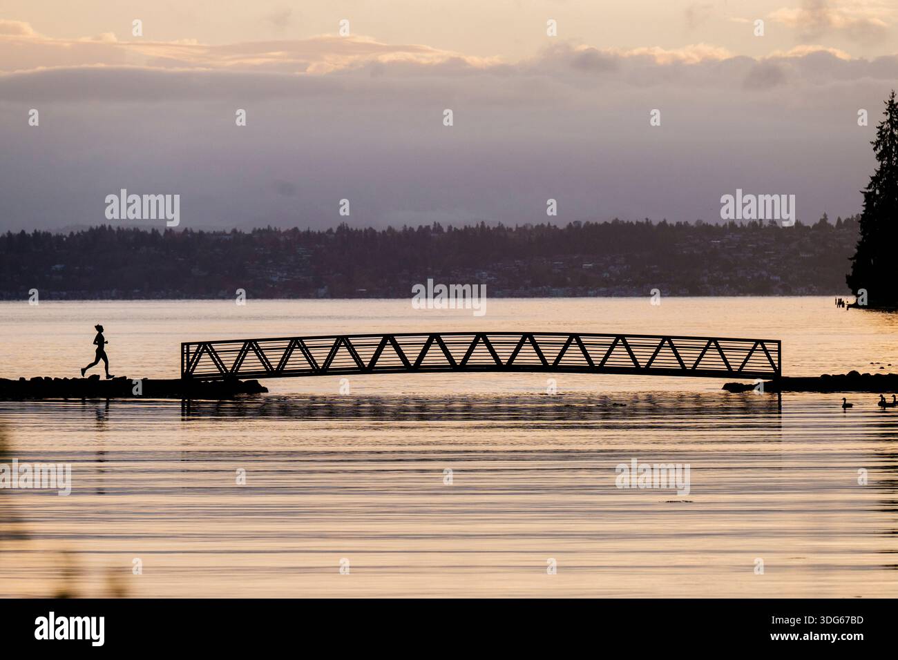 A person jogs along a bridge at sunset over a calm body of water with ...