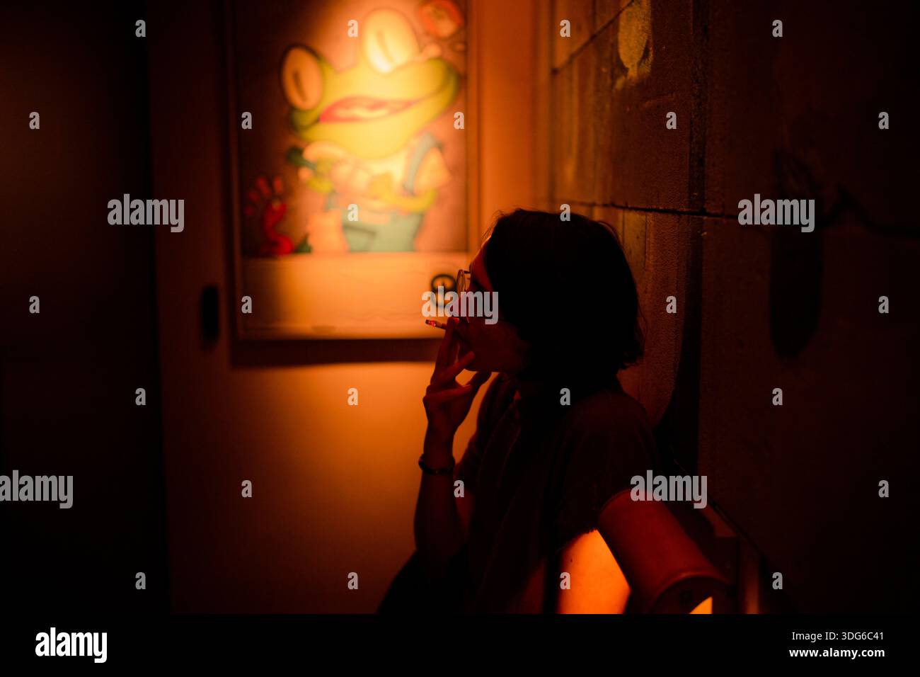 Woman smoking in a dimly lit room with a frog poster on the wall ...