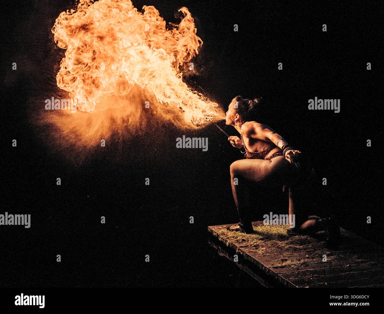 Fire performer kneeling on wooden platform, breathing a dramatic burst ...