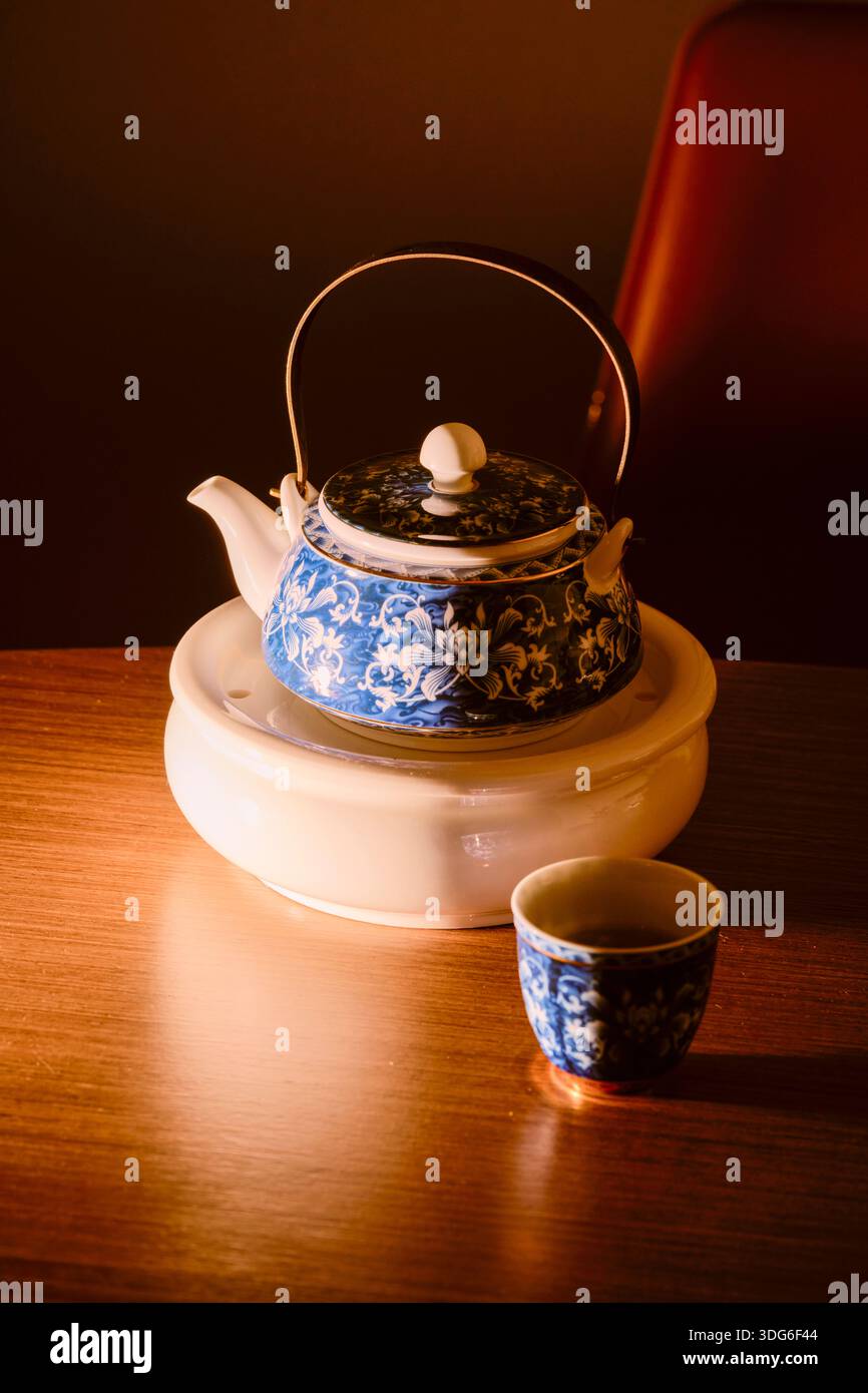 Blue and white ornate teapot with a teacup on a wooden table lit warmly ...