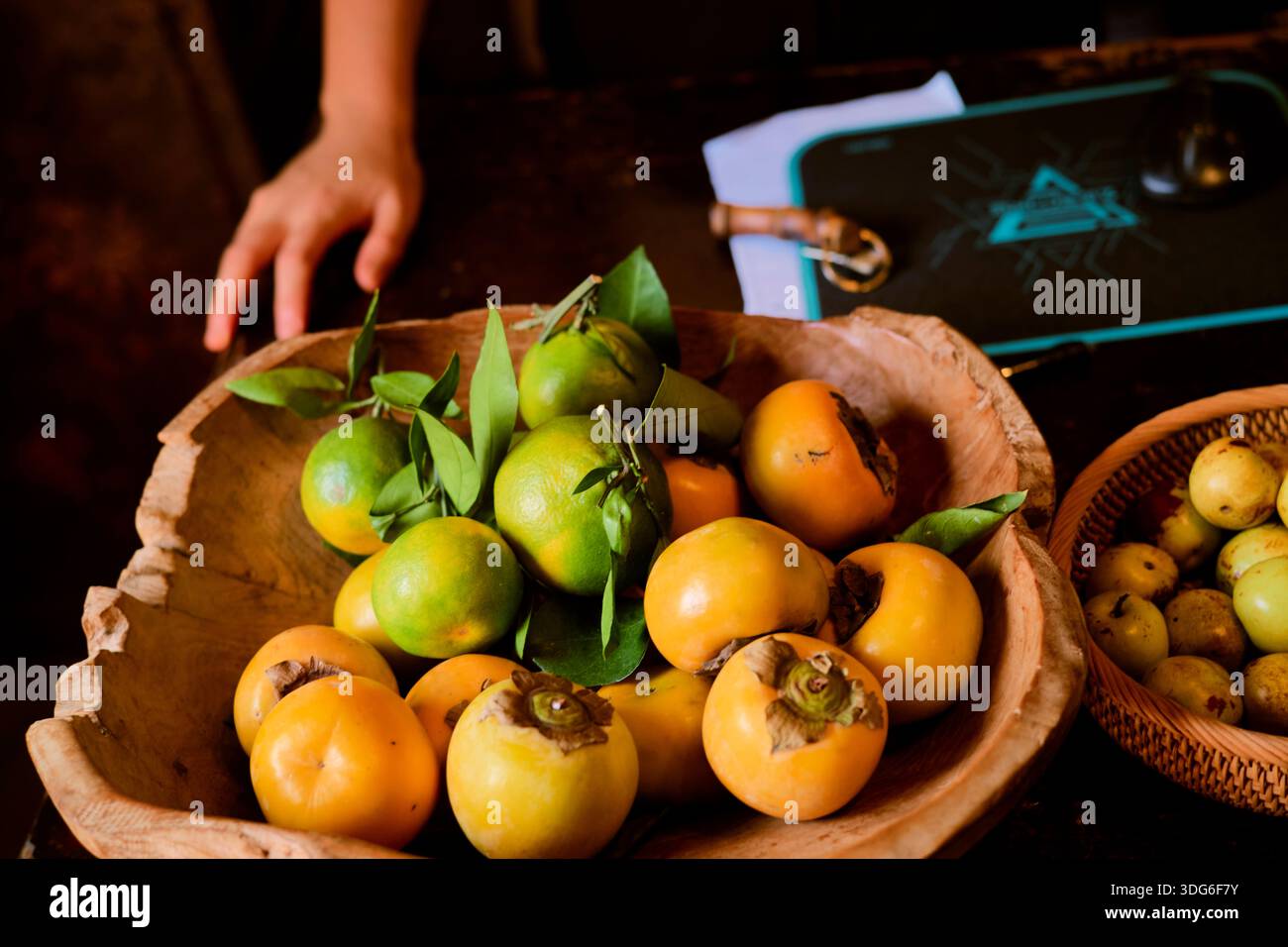 A wooden bowl with fresh persimmons and limes on a dark table, with a ...