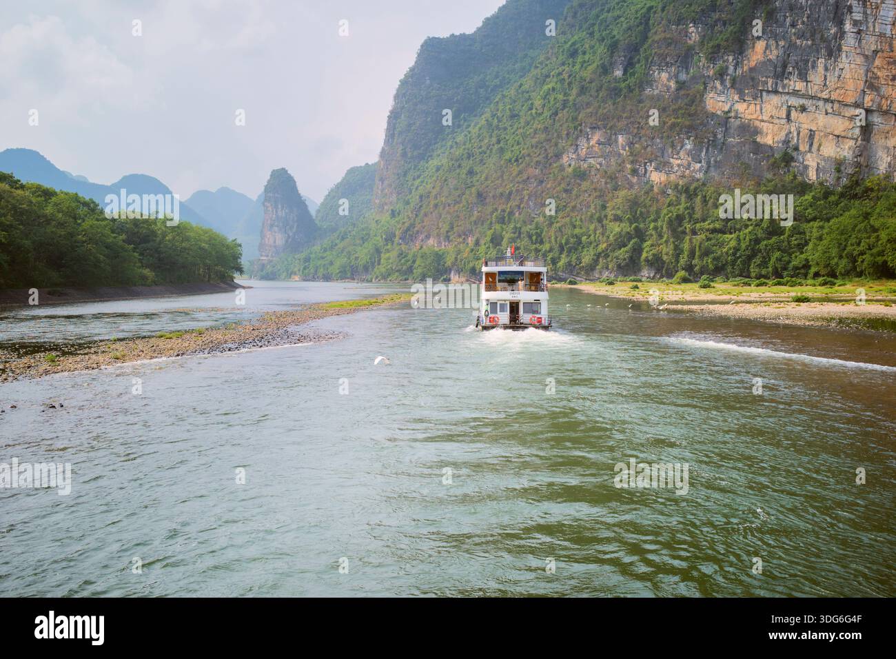 A boat cruises down a river flanked by lush green mountains and rocky ...