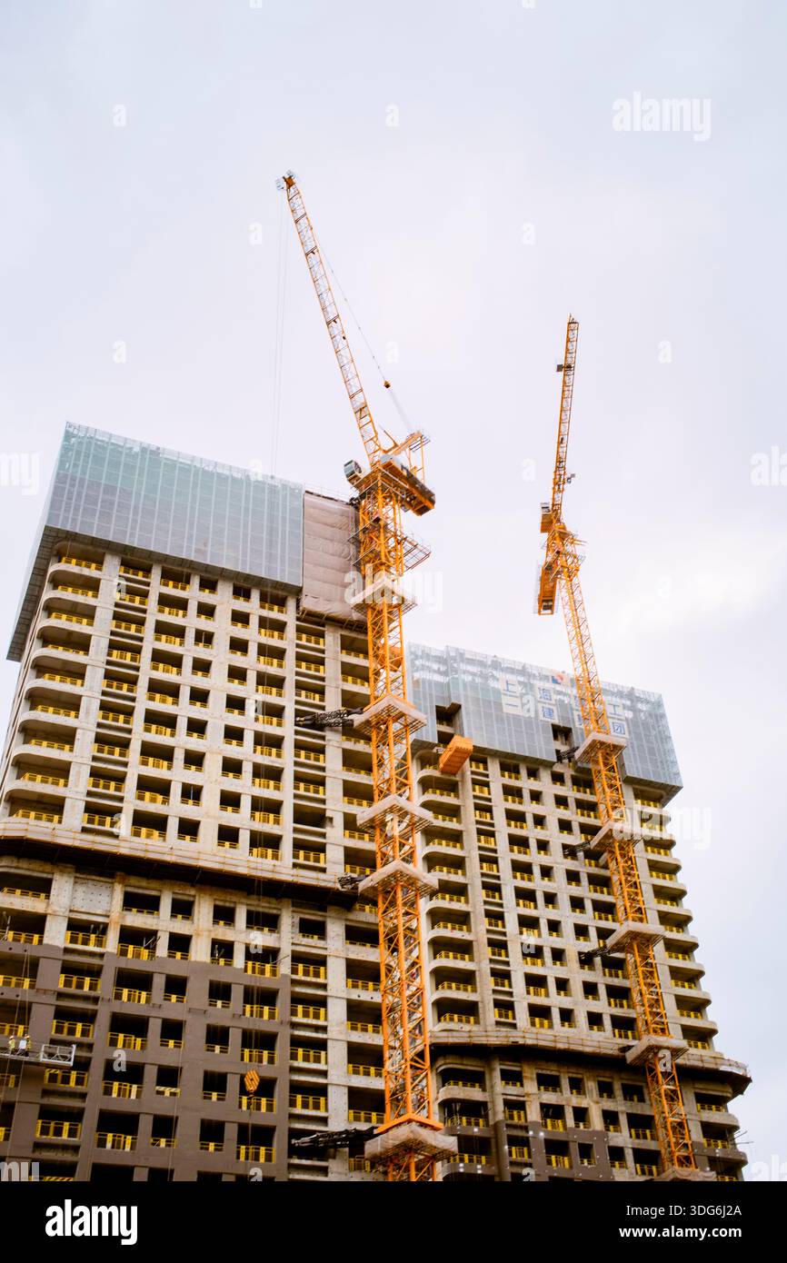 Tall construction cranes beside a high-rise building under a cloudy sky ...