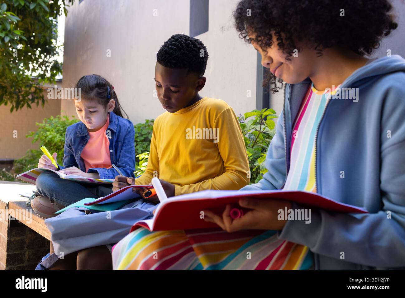Diverse children sitting on low stone bench in courtyard with backpacks ...