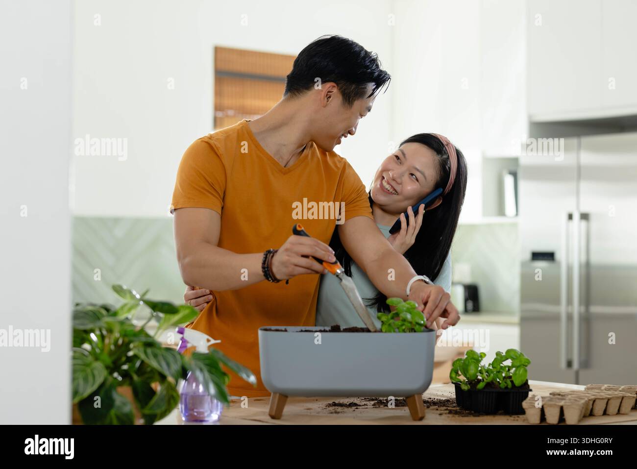 Asian couple potting herbs at kitchen counter with grey planter using ...