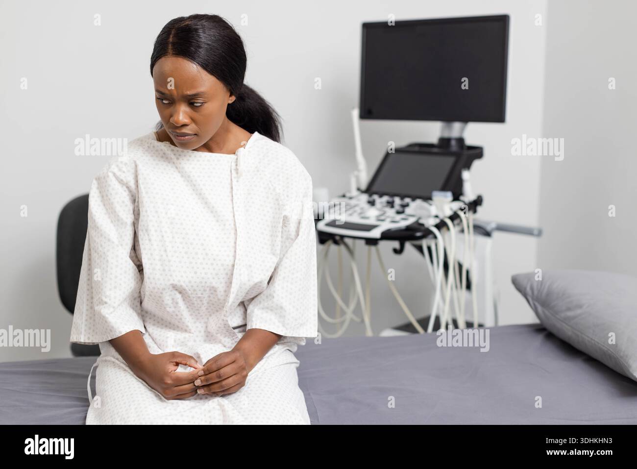 Female patient wearing hospital gown sitting on exam bed clasping hands ...