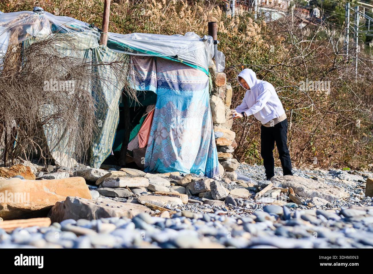 Individual in white hoodie touches rock foundation of tarp-draped hut ...