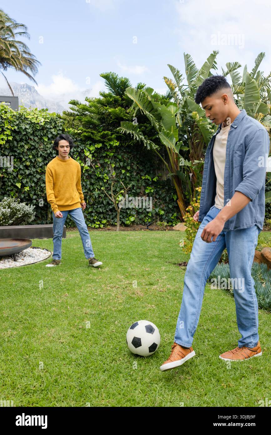 Diverse male friends kicking soccer ball on backyard lawn near fire pit ...