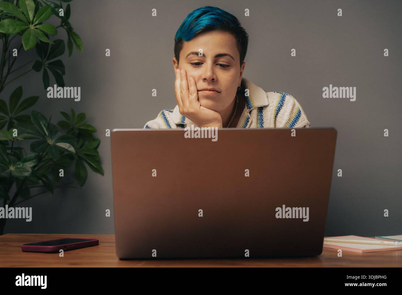 A young non-binary person with blue hair sits at a wooden desk, looking ...