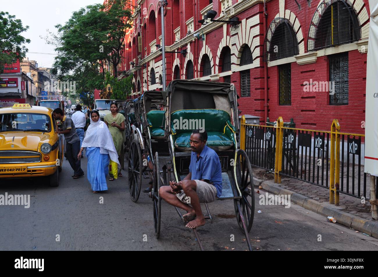 Hand-pulled rickshaw driver waits along a street beside a red colonial ...