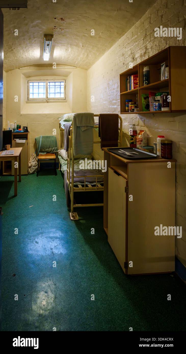 Small austere room with single bed, desk and shelves, barred window and ...