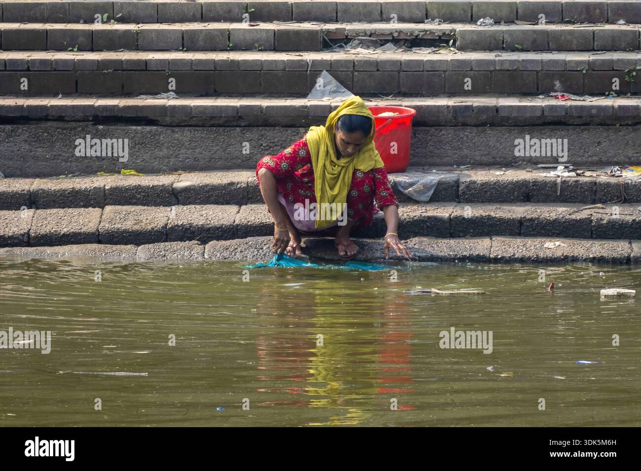 A South Asian woman washing clothes on a stone ghat by a rural pond ...