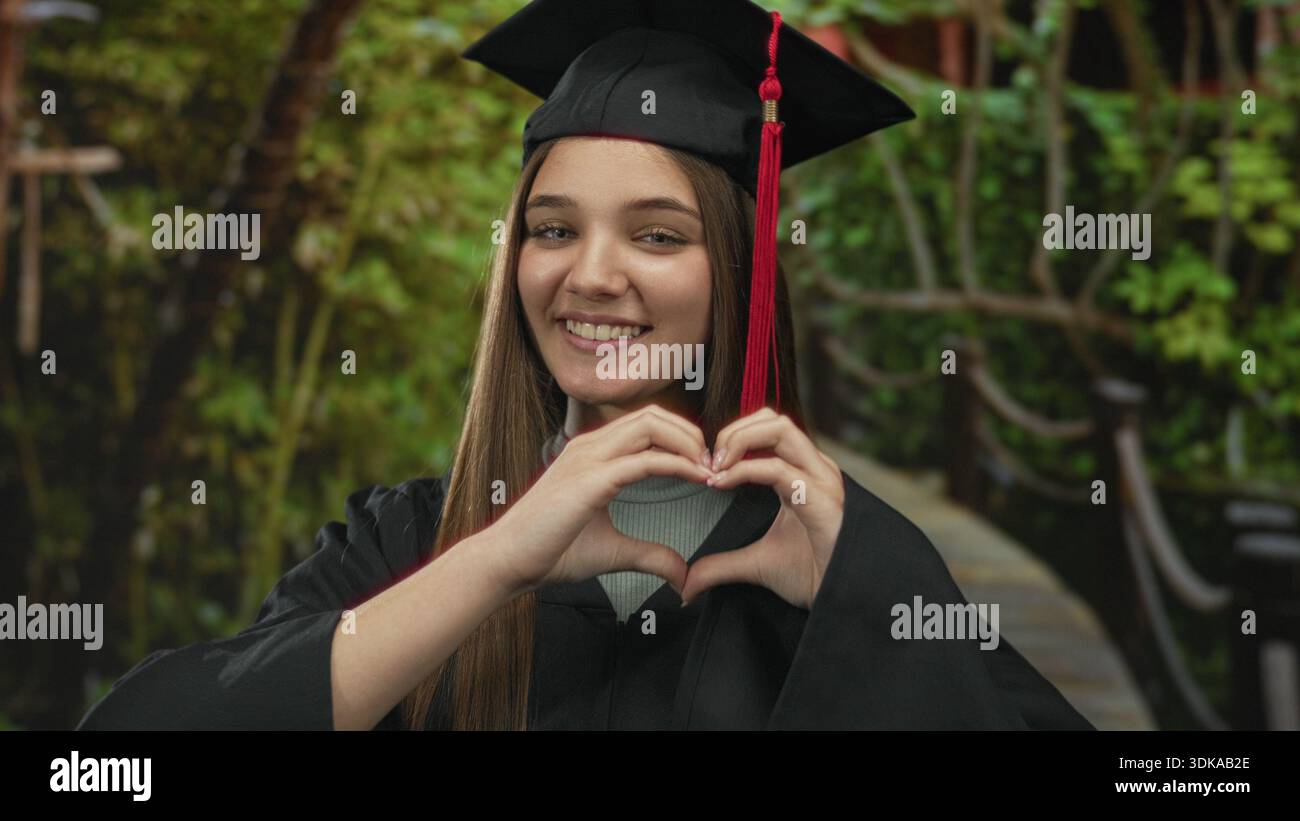 Woman wearing graduation cap and gown forming heart with hands in green ...
