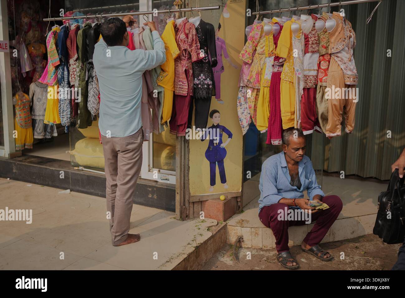 A vegetable vendor counts money while a shopkeeper sets up dresses for ...
