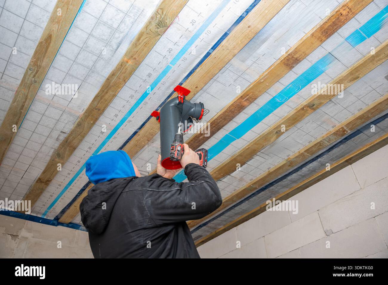 A worker in a blue hood and black jacket uses a power drill to install ...