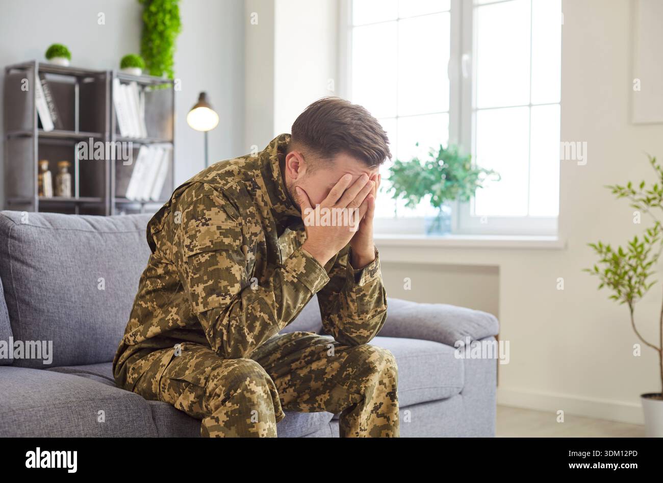 Young man in military camouflage uniform sits on couch expressing deep ...