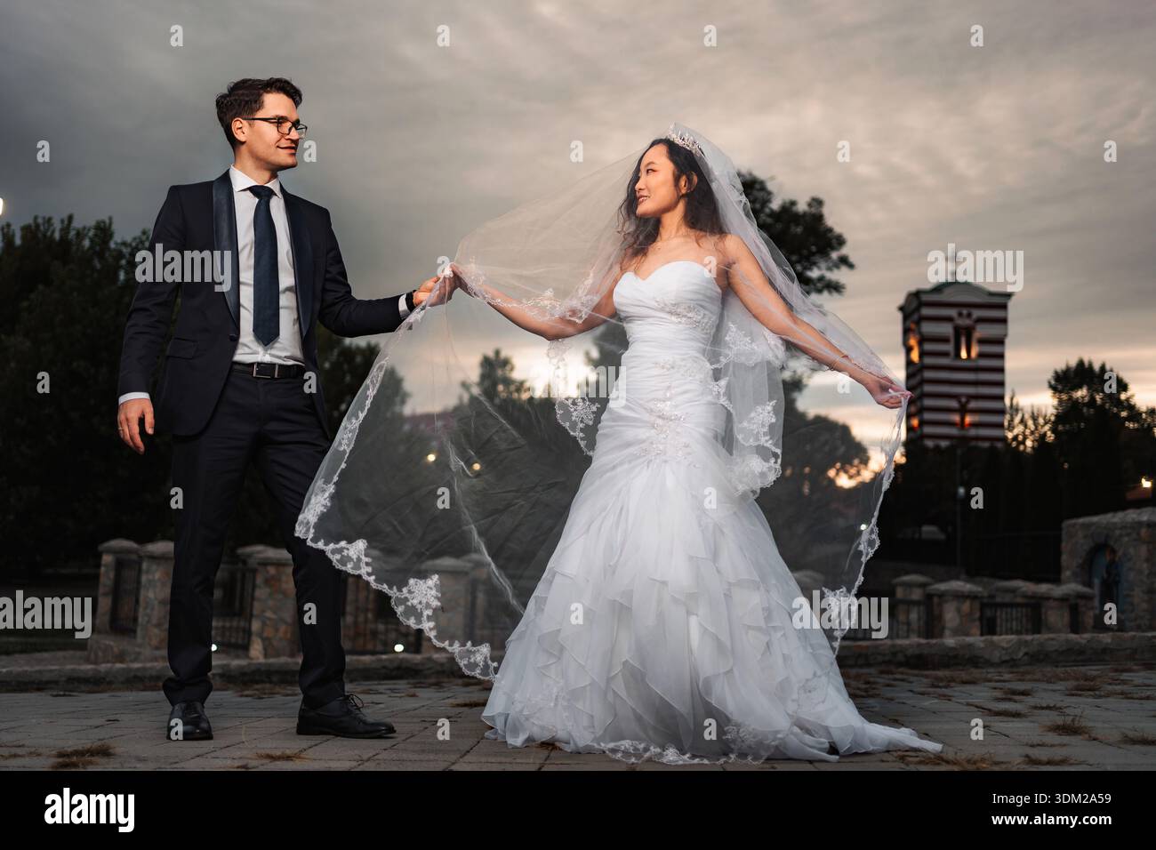 Groom holding bride's flowing veil outdoors at dusk, celebrating their ...