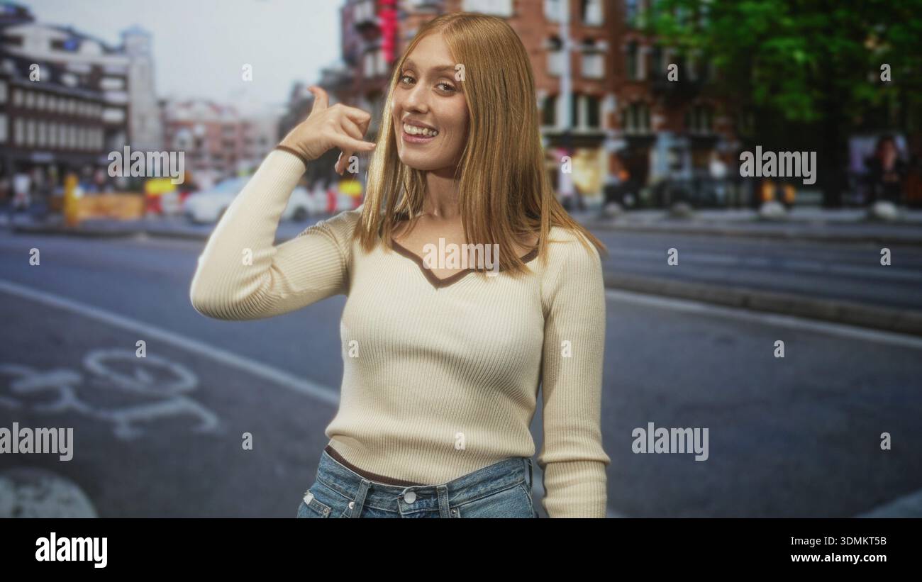 Woman redhead smiling and making a call hand gesture on a city street ...