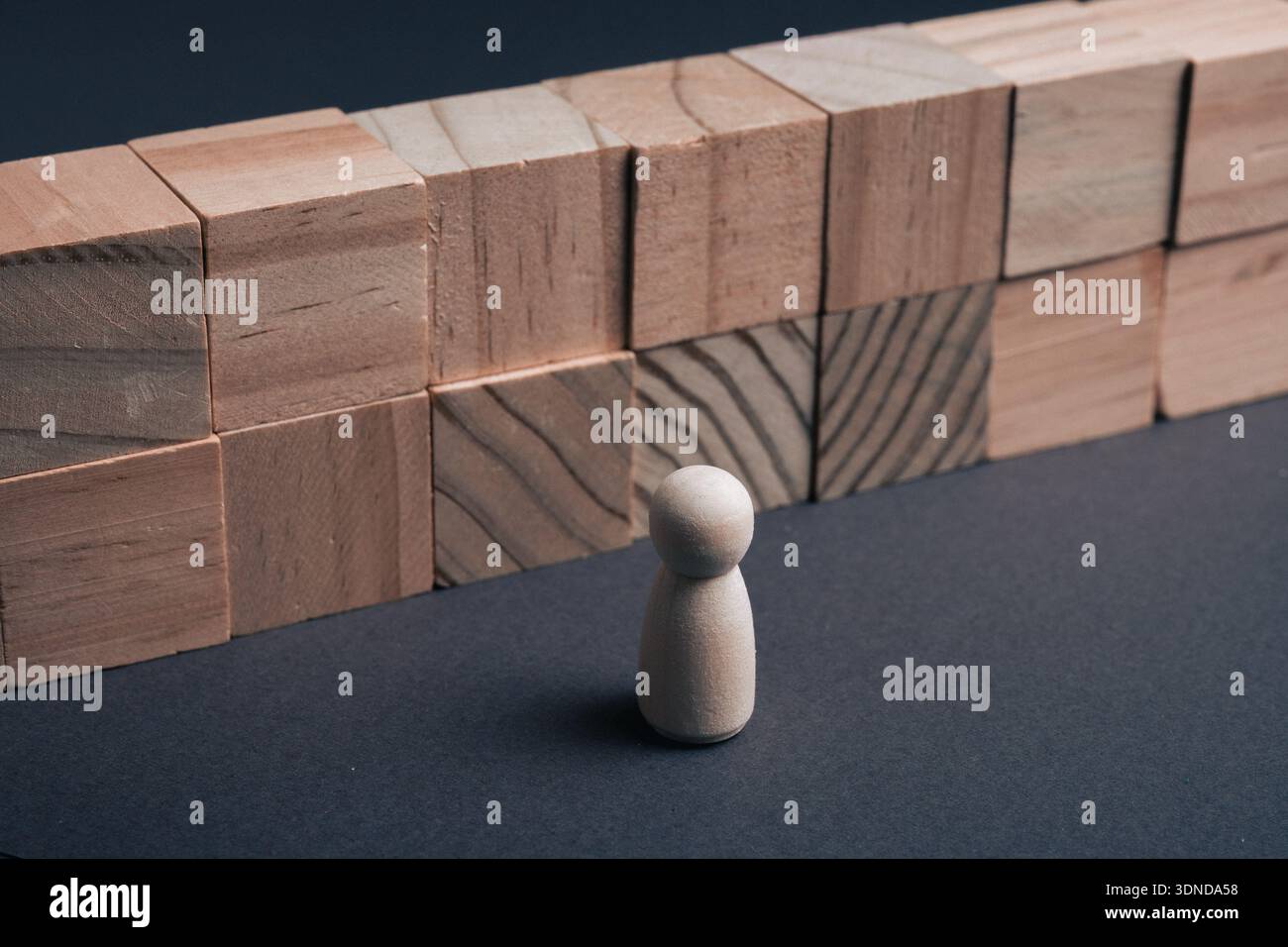 A wooden peg figure stands alone facing a tall wall constructed from ...