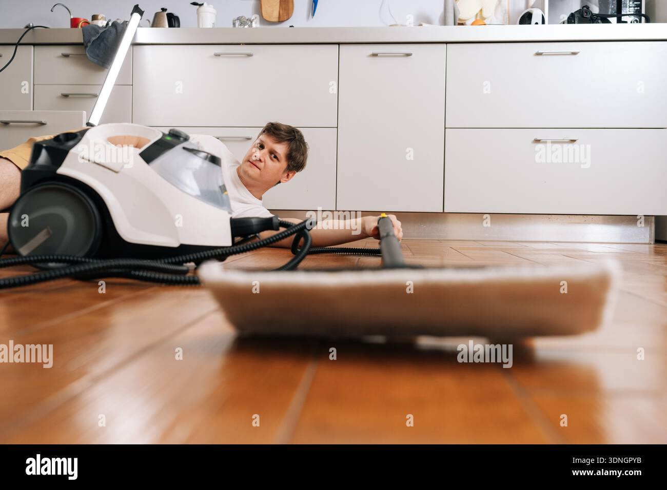 Man lying on kitchen floor using extended-handle steam cleaner to deep ...