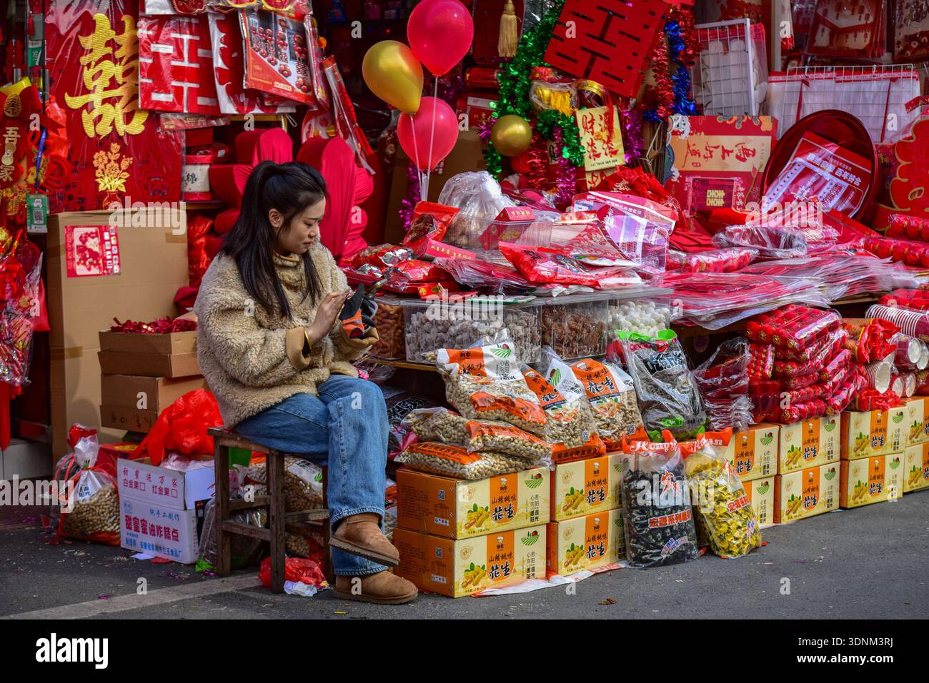 A young woman sits on a stool and uses her phone at a stall in the ...