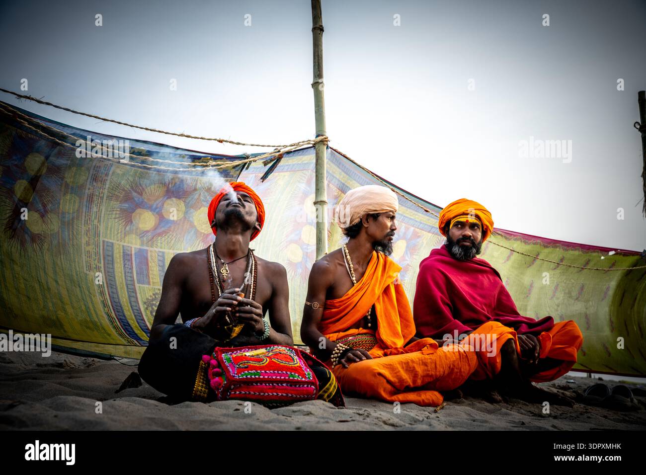 Three Indian sadhu men in traditional attire sitting on the ground, one ...