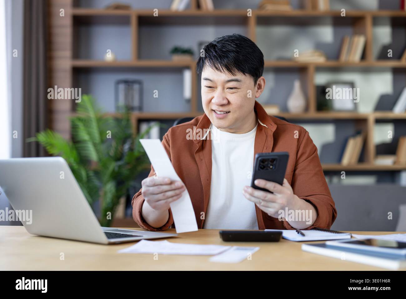 Asian man smiling at home office desk, holding a receipt and using ...