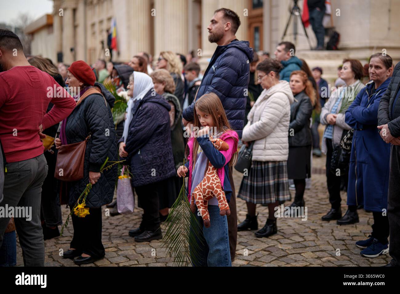 A child holding a toy giraffe eyes the camera while attending an ...