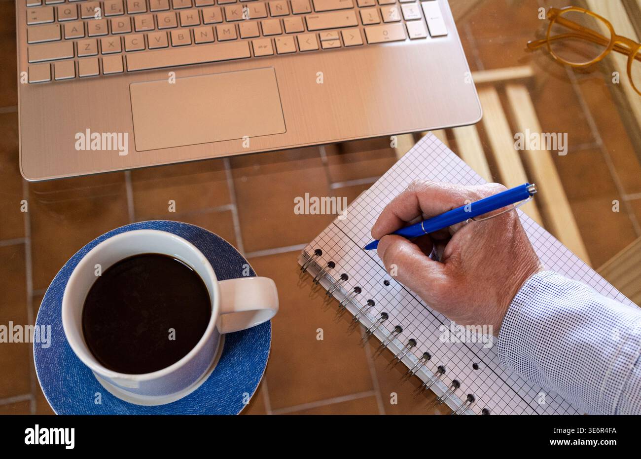 Person's hand making notes in a notebook on a glass table with laptop ...